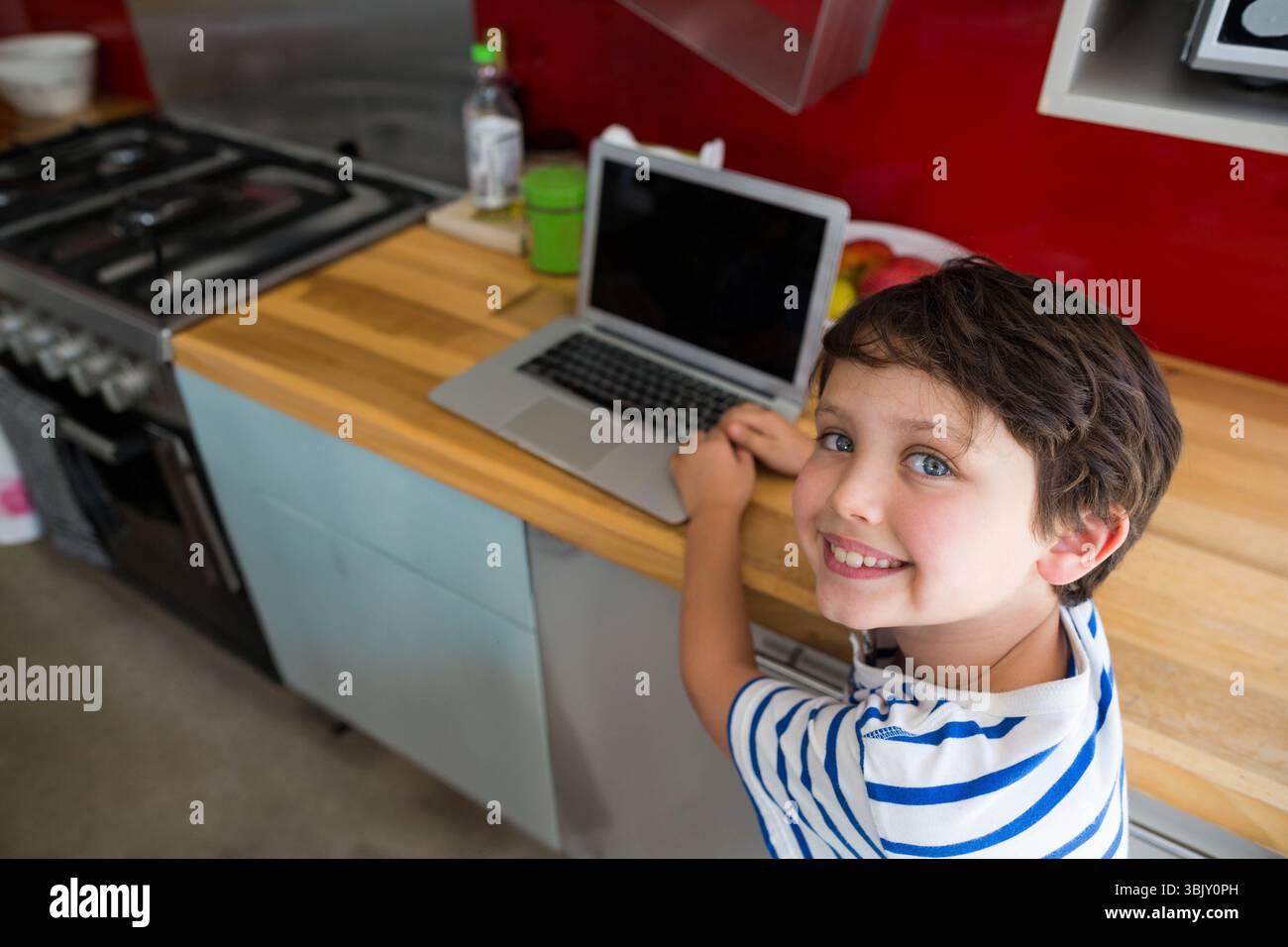 School-aged boy standing at wooden kitchen counter typing on laptop ...