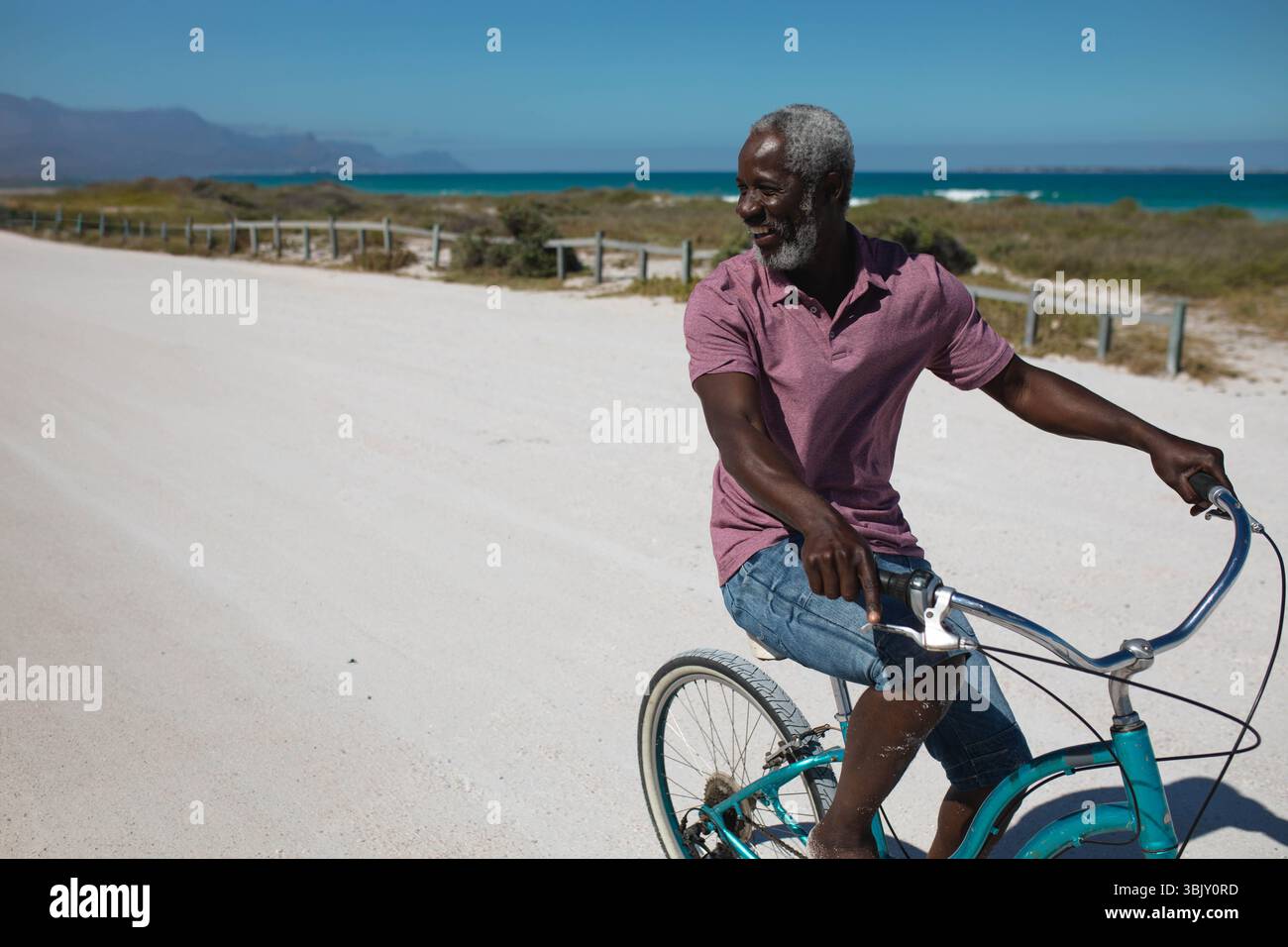 Senior African American man riding teal cruiser bike along coastal path ...