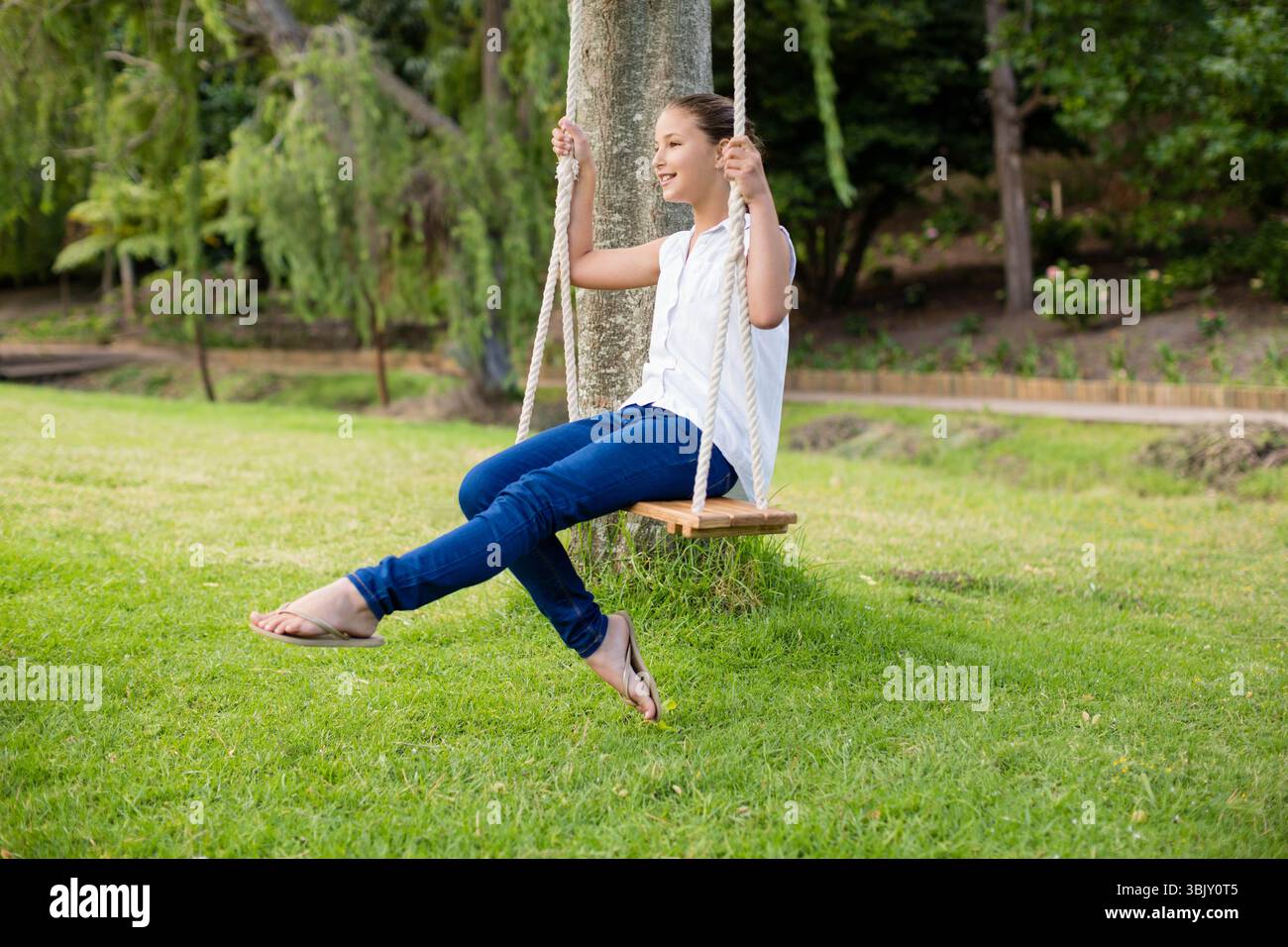 Preteen girl swinging on wooden plank swing in backyard with thick ropes and sturdy tree trunk ...