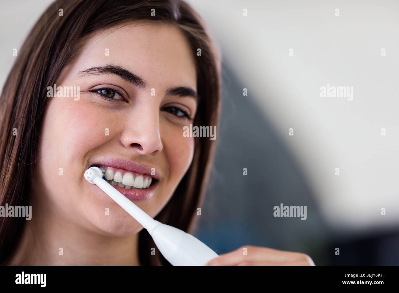 Woman brushing teeth with white electric toothbrush in bathroom near mirror wearing light top ...