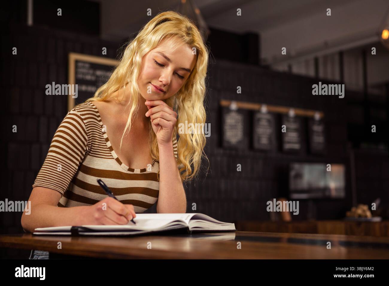 Blonde woman writing in open notebook at café table under lights by ...
