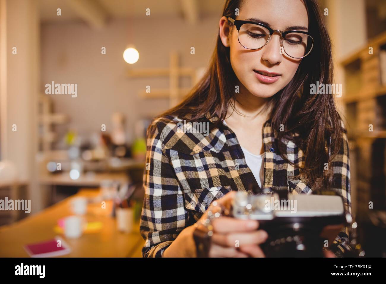 Woman wearing eyeglasses inspecting vintage camera in office with coffee mugs and notebooks ...