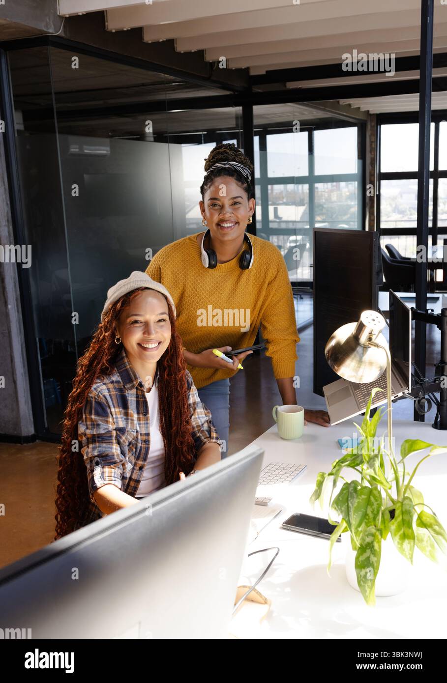 Diverse women collaborating in modern office, smiling and using computer for coding project ...