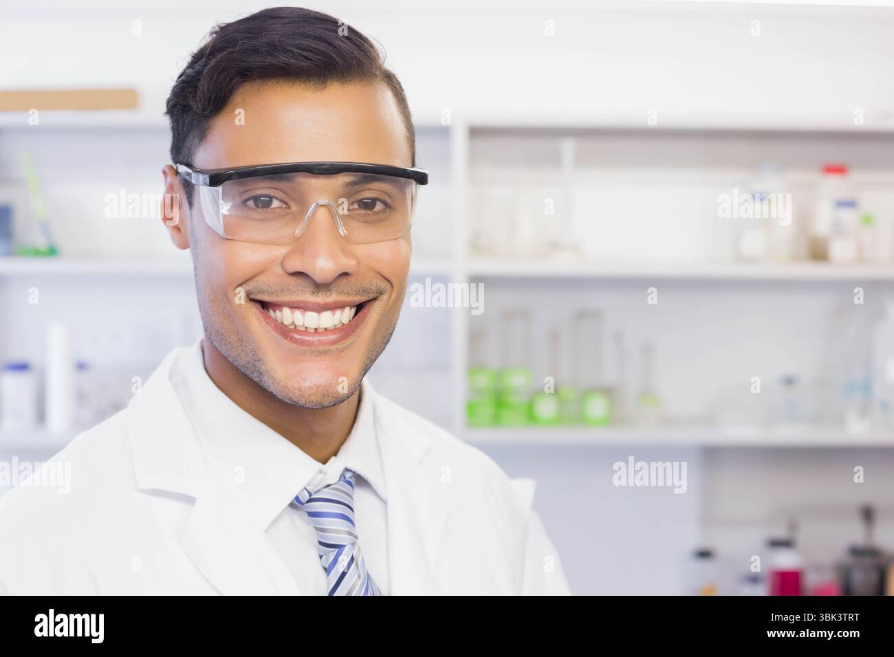 Smiling male scientist in safety goggles, lab coat, striped tie posing in lab with green ...