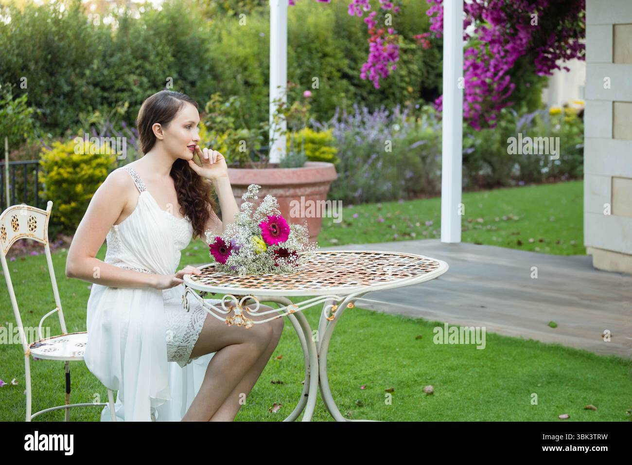 Woman wearing white dress sitting garden patio by iron table holding ...