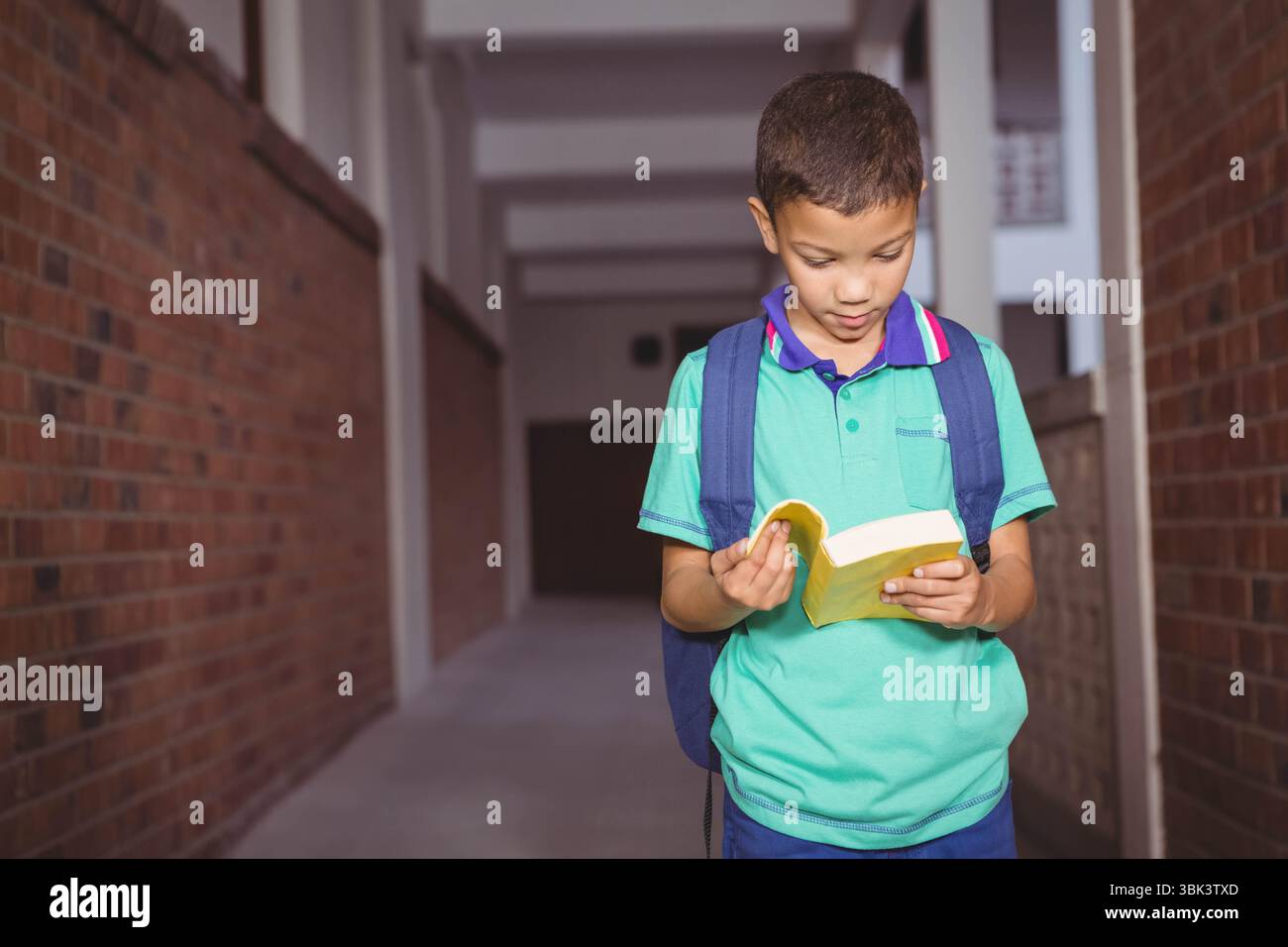 Boy standing in school corridor with brick walls reading yellow book ...