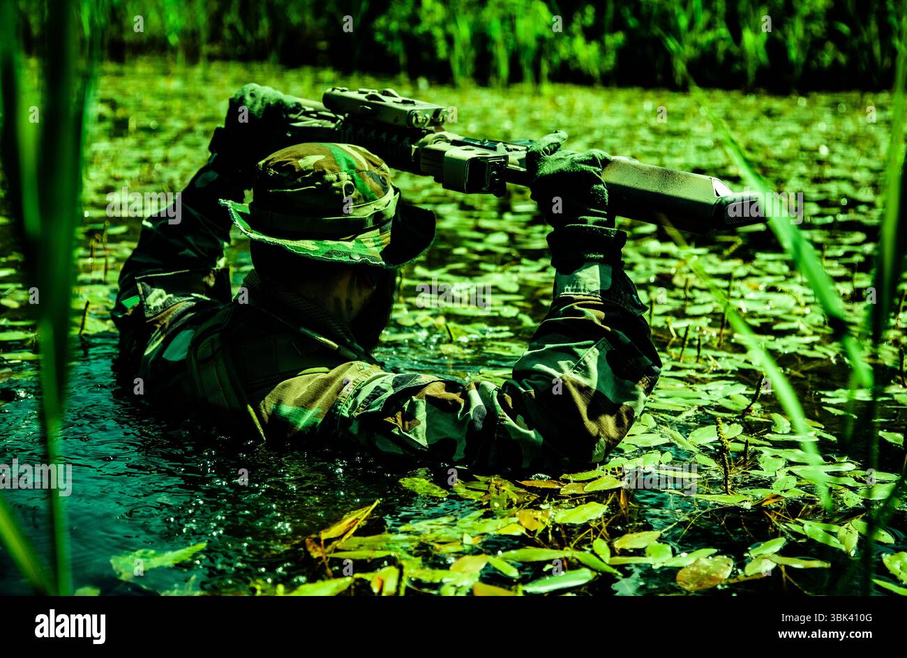 Soldier in camouflage wades through a swamp holding a rifle above the ...