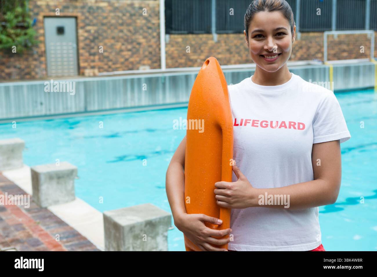 Female lifeguard standing at pool edge holding orange rescue buoy at ...