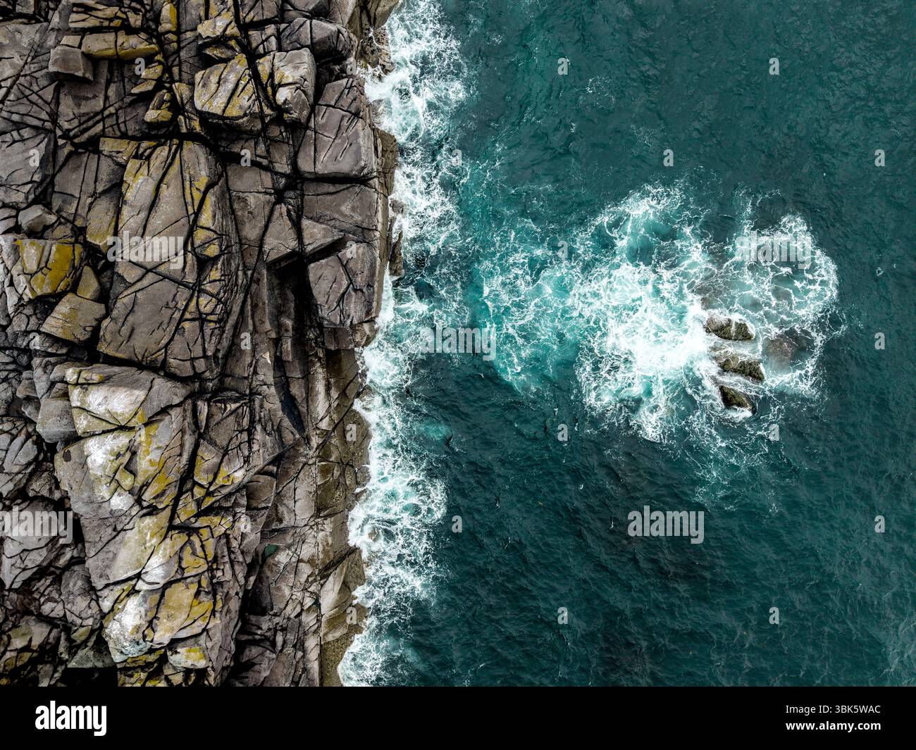 Aerial view directly above rocky cliffs into rough seas in a coastal ...