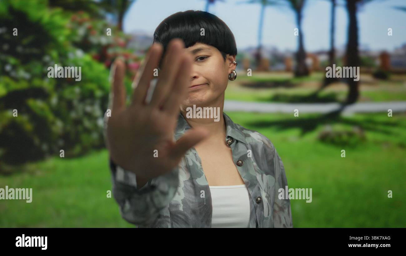 Young chinese woman making a stop gesture with her palm in an outdoor park setting, surrounded ...