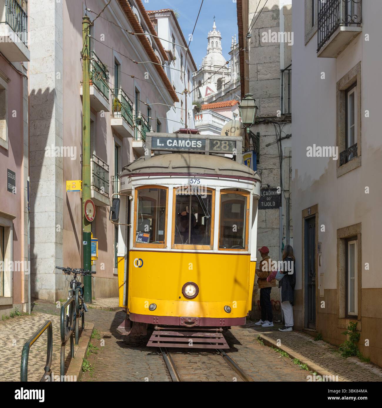 A vibrant yellow Tram 28 approaches on a narrow Lisbon street, with ...