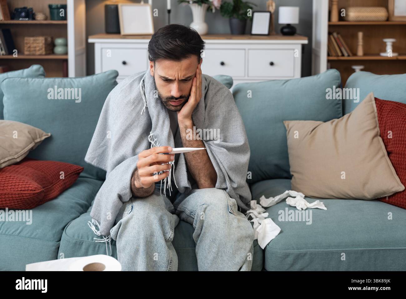 Ill man lying wrapped in blanket and checking temperature at home ...