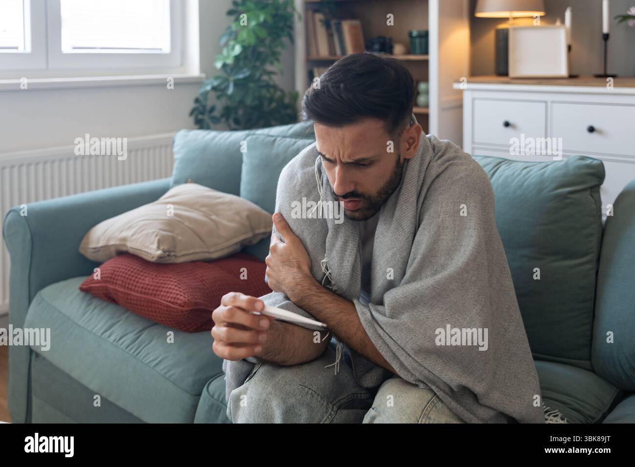 Ill man lying wrapped in blanket and checking temperature at home ...