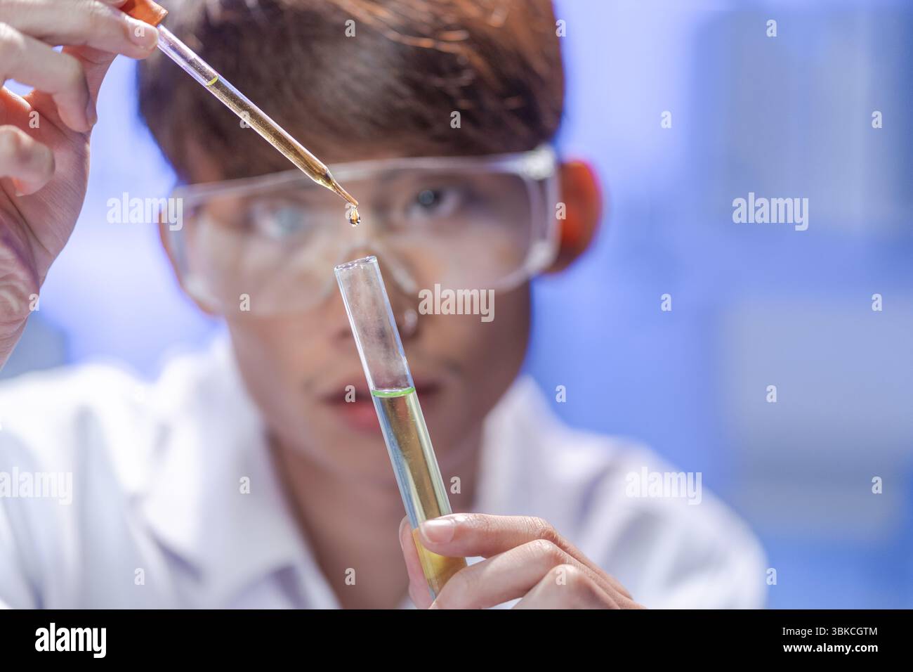 A young male researcher in a lab coat and safety goggles carefully adds ...