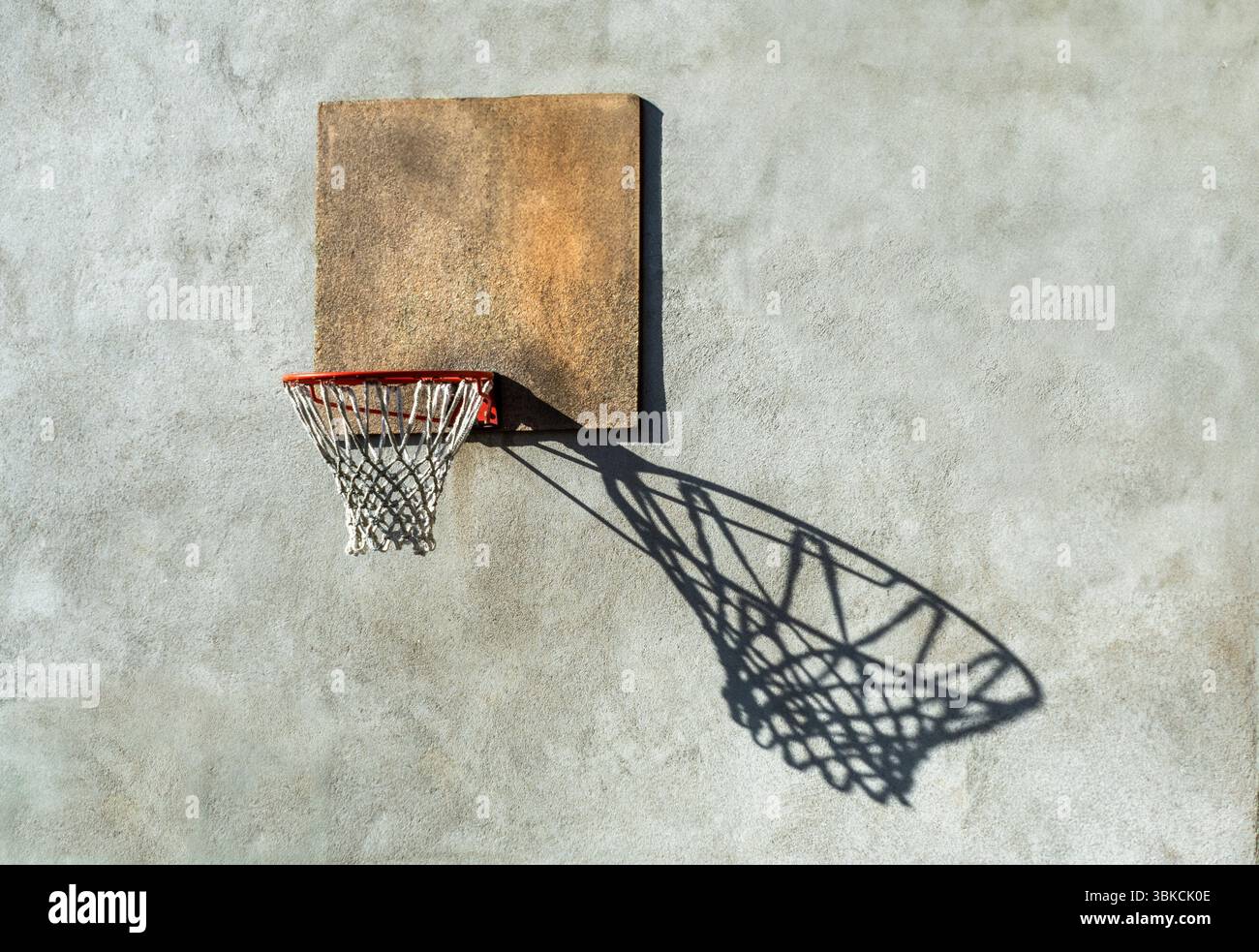 A worn basketball hoop is mounted on a textured wall, with the sunlight ...