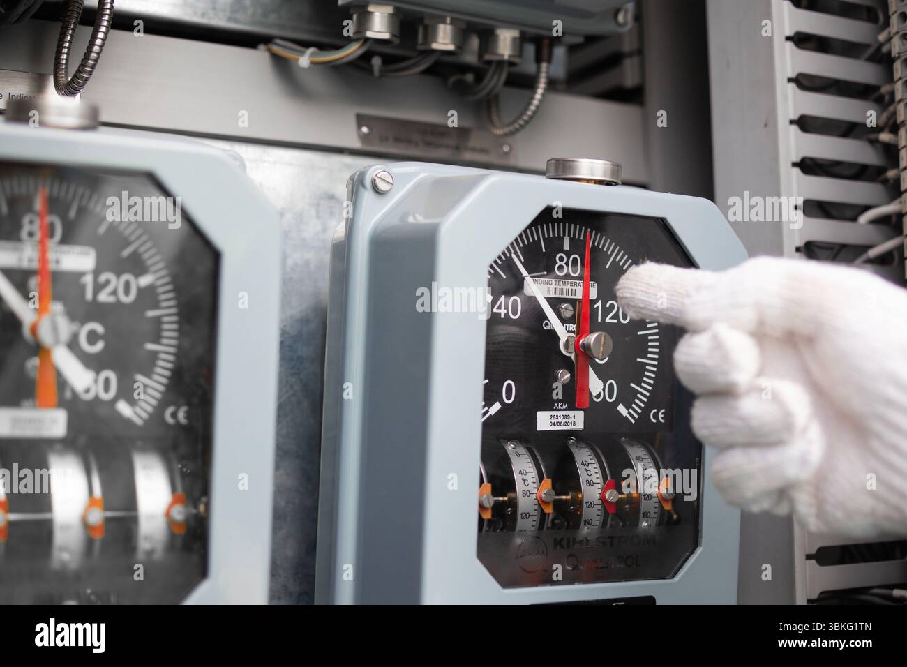 A gloved hand points to temperature gauge inside power transformer control panel at substation ...