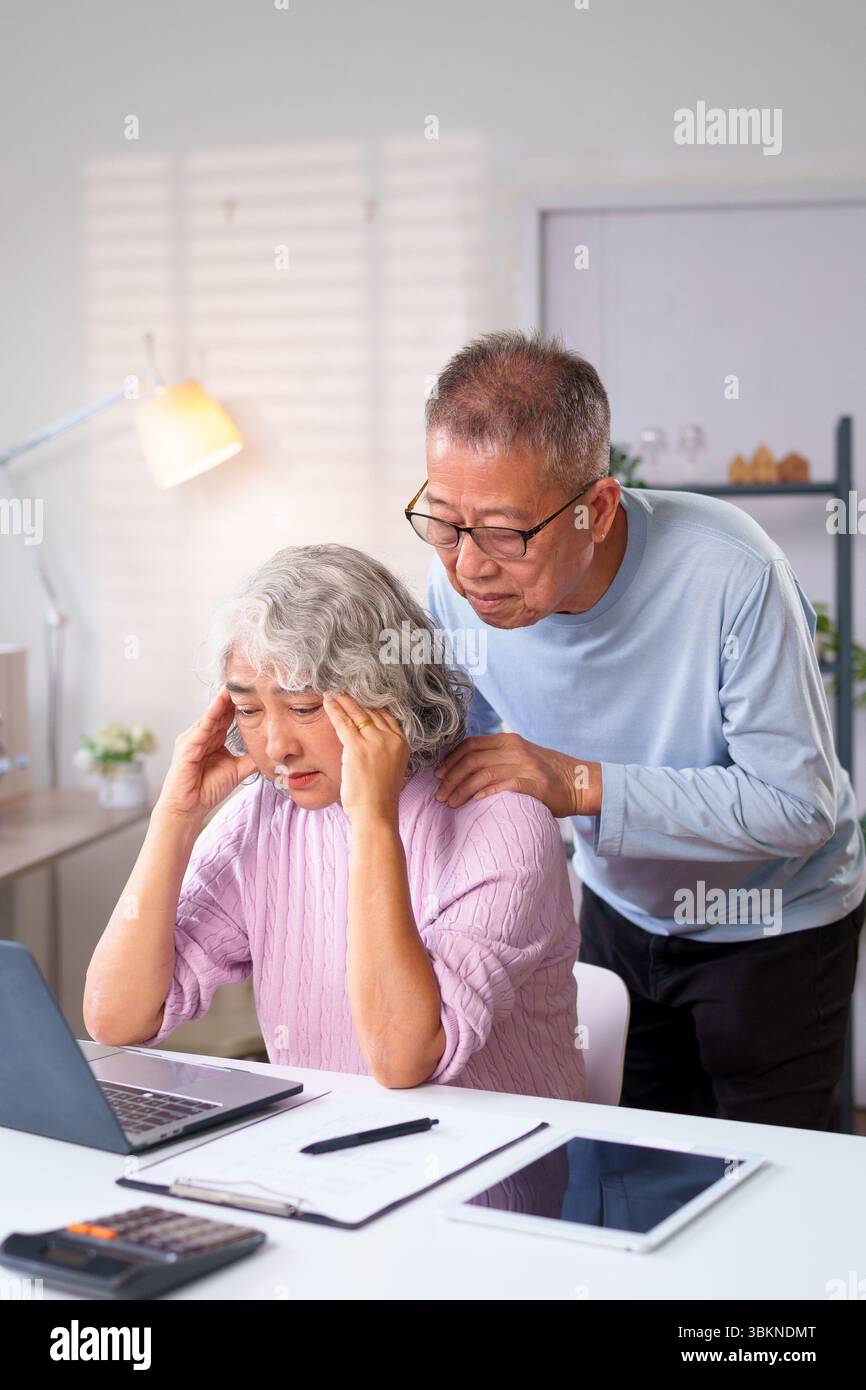 An older Asian woman experiences stress at her home desk, her loving ...