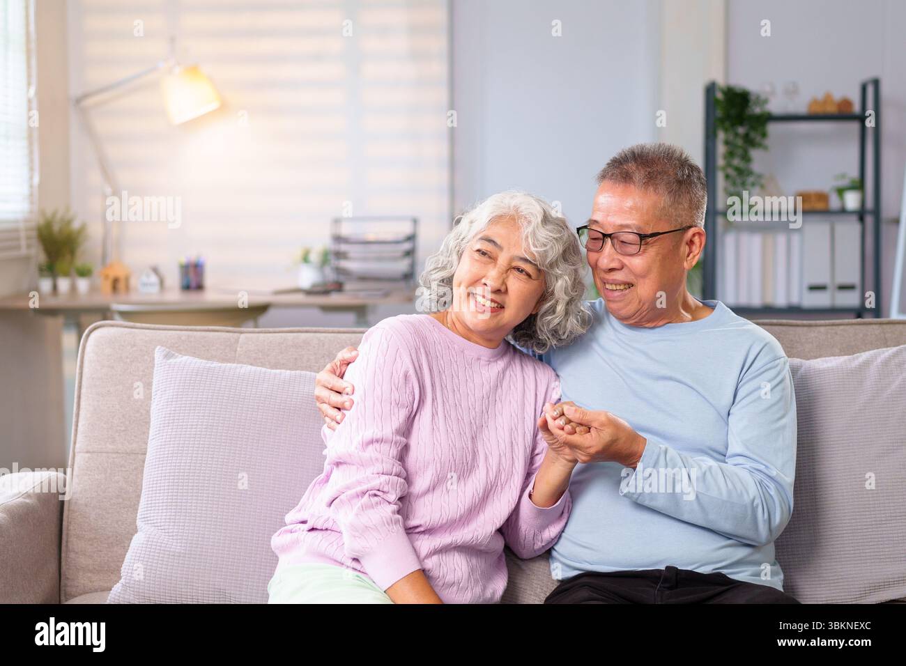An older Asian couple enjoys a peaceful moment on their sofa expressing deep love and care. This ...