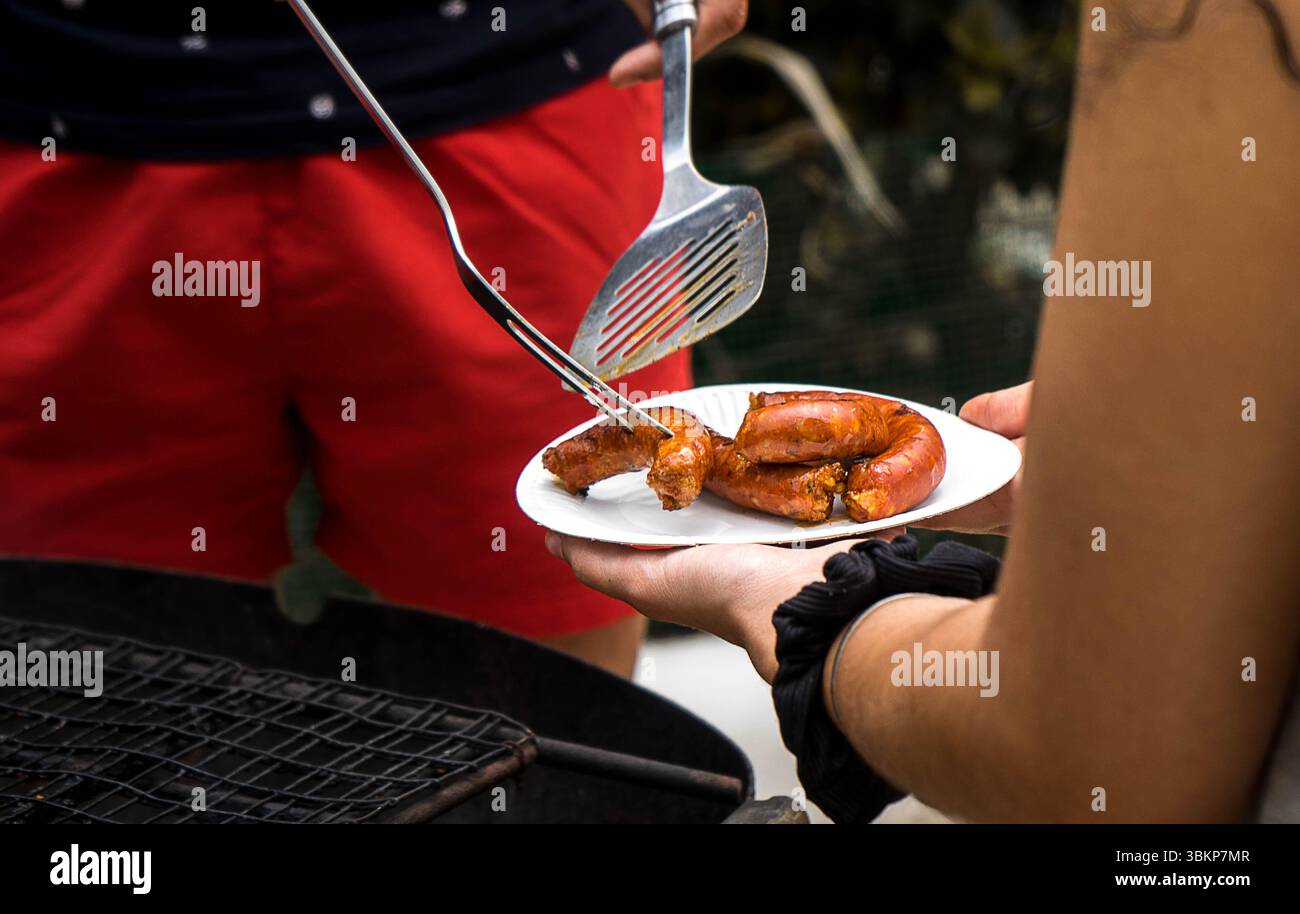 A person is serving sausages from a barbecue onto a plate, with another ...