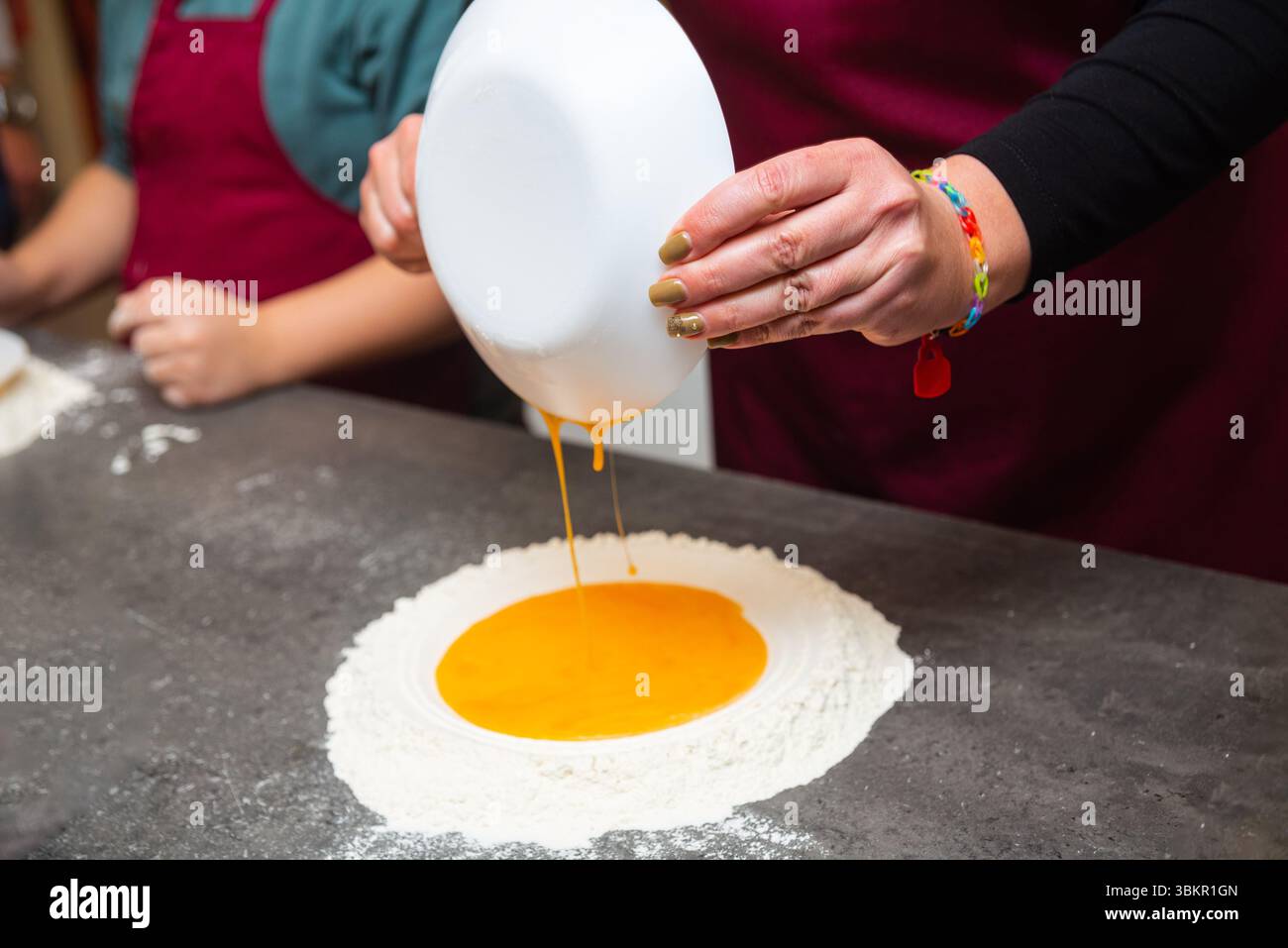 A person pours beaten eggs into a flour well on a countertop, beginning ...