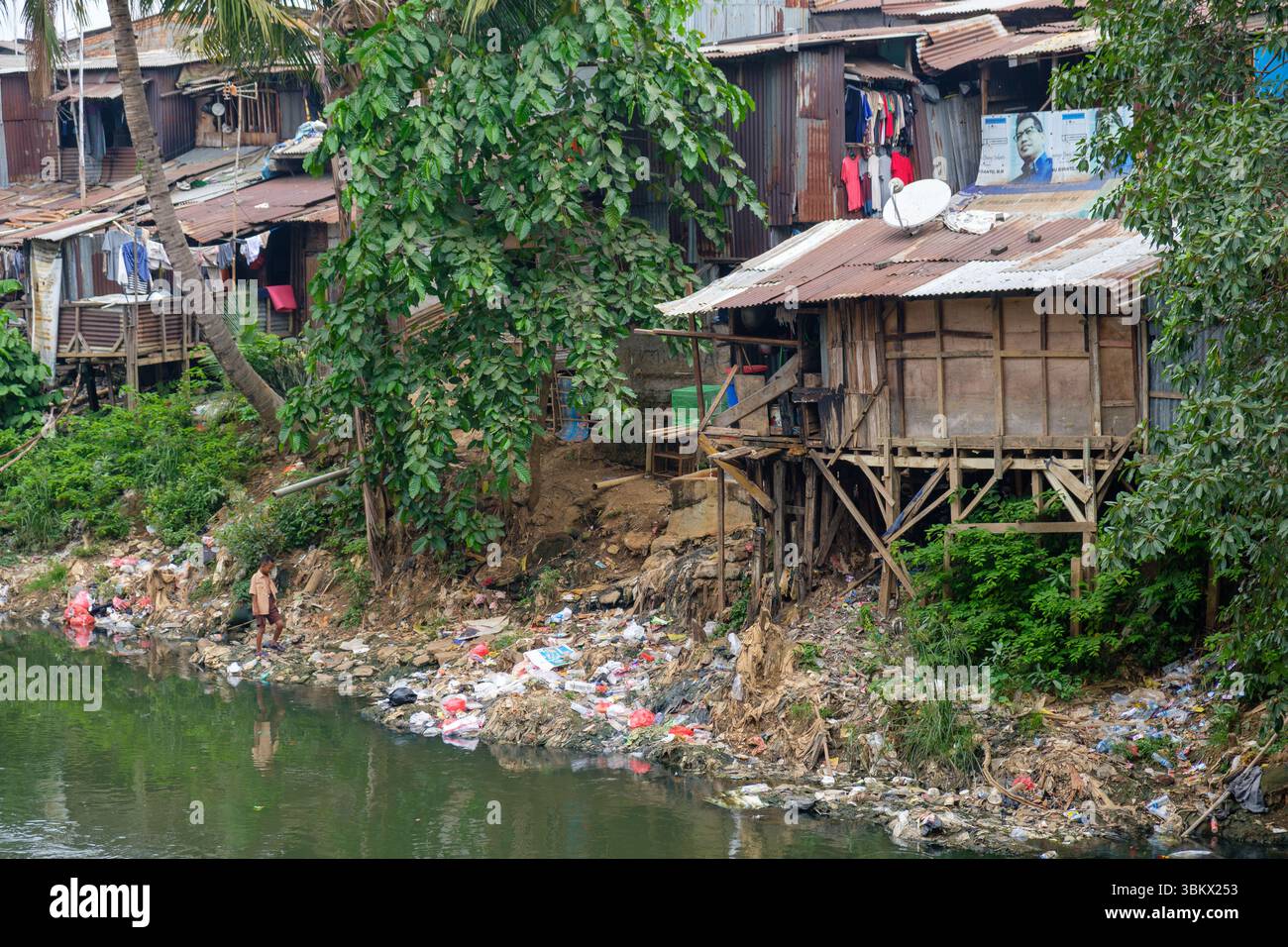 A person stands by a riverbank covered in trash next to slum houses ...