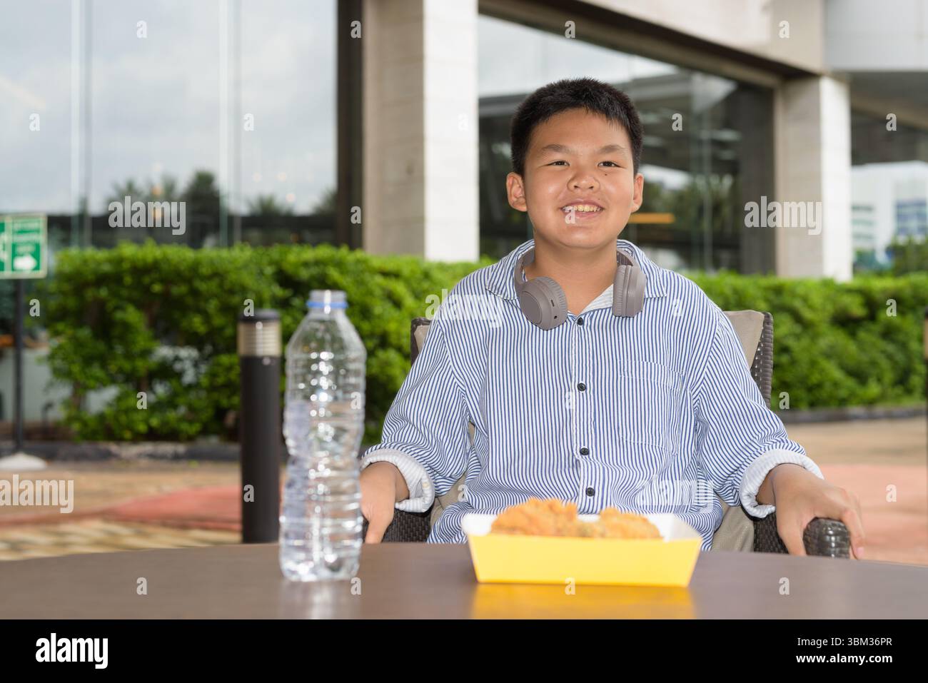 Thai student boy eating fried chicken with headphones at outdoor table ...