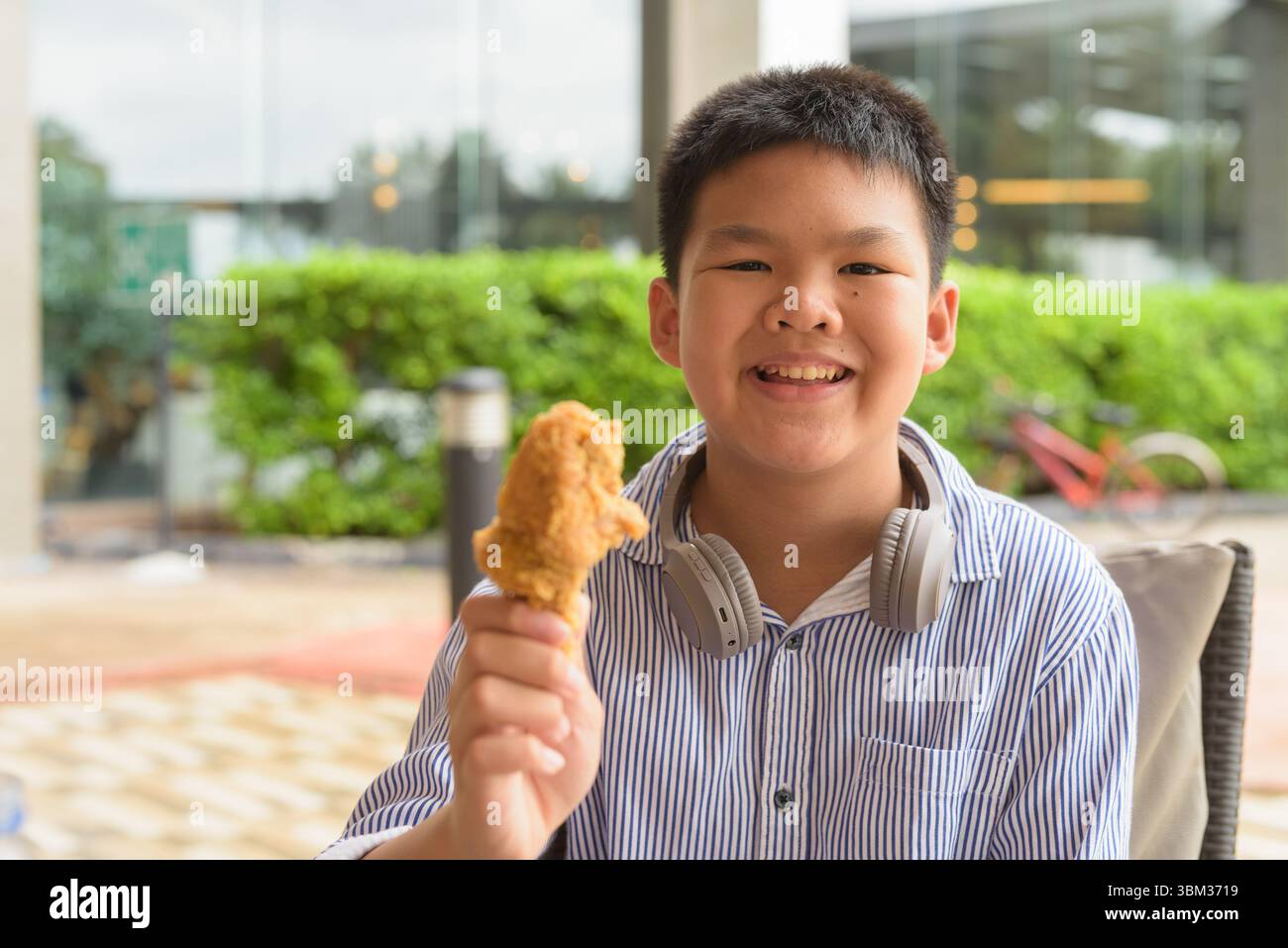 Thai student boy eating fried chicken with headphones at outdoor table ...