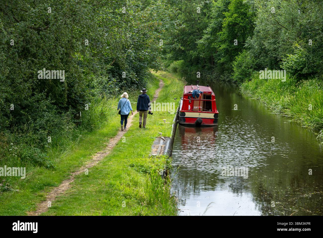 Two people walk along a towpath past a red narrowboat moored on the ...