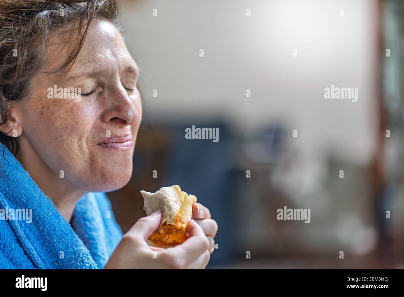A woman is eating a sandwich and smiling. She is wearing a blue robe ...