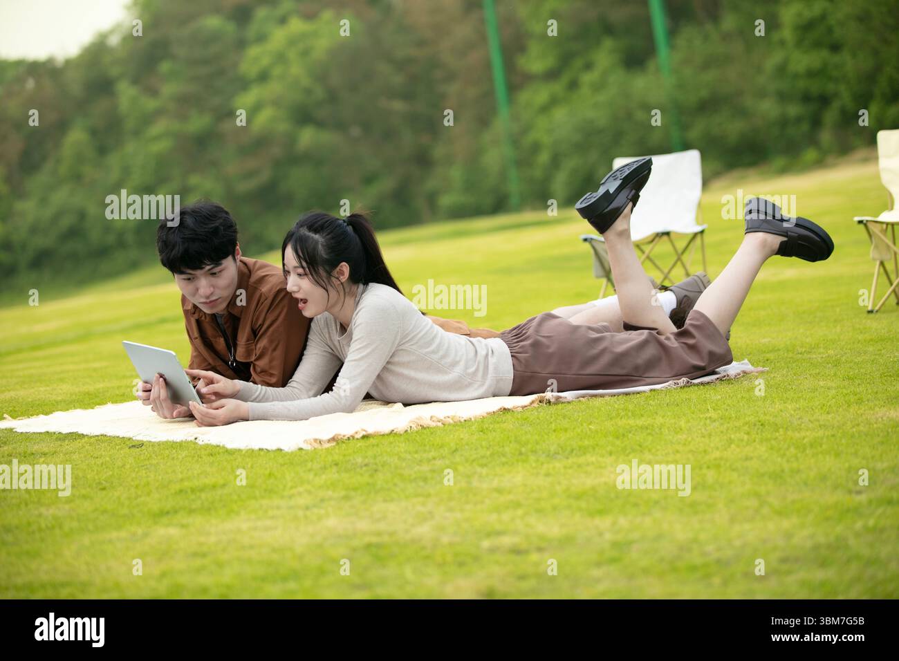 A young couple lying side by side on an outdoor mat watching a tablet ...