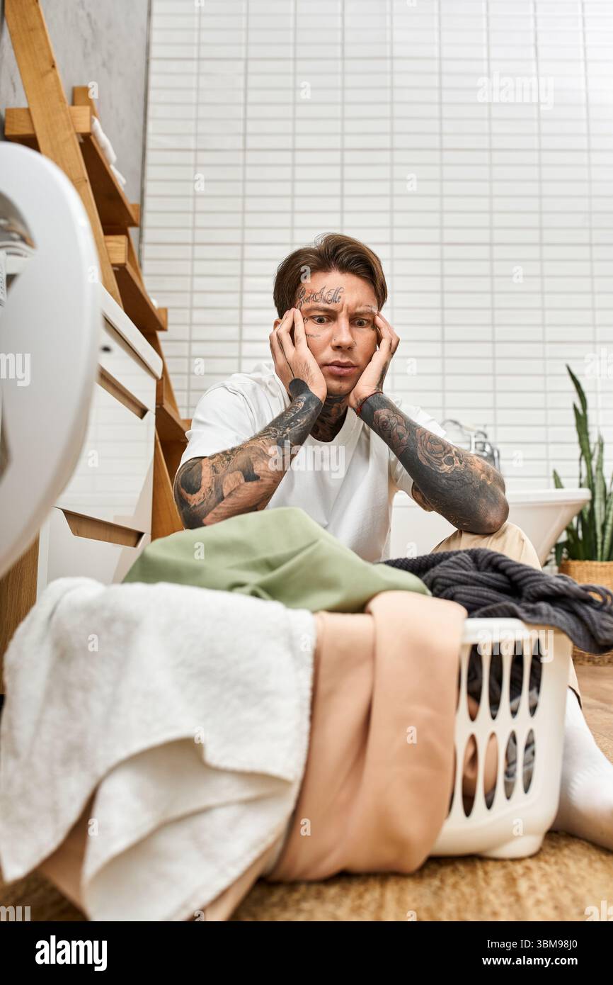Handsome young man with tattoos sits amid laundry piles, reflecting on ...