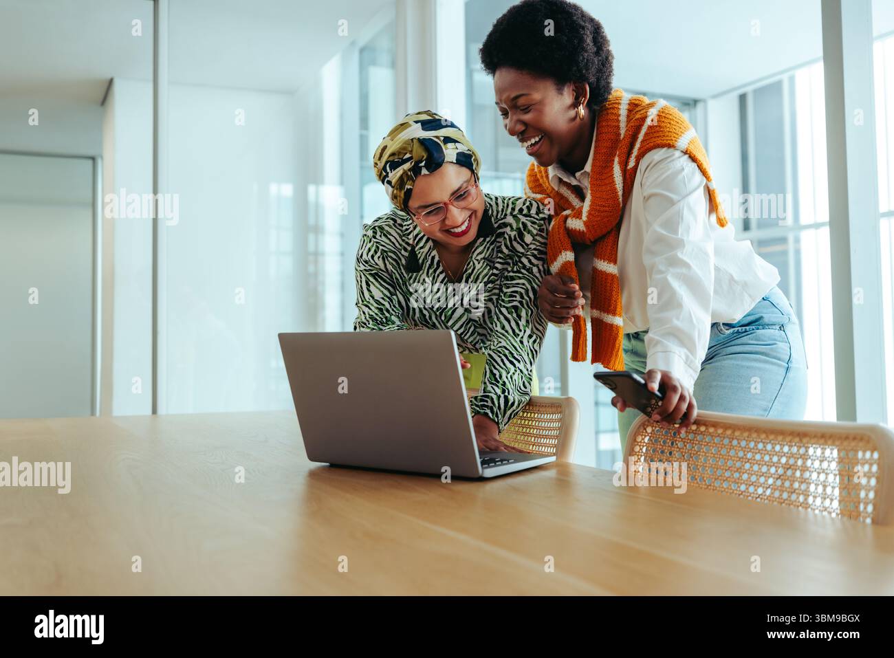 Two diverse women are joyfully collaborating in a stylish office environment, using a laptop to ...
