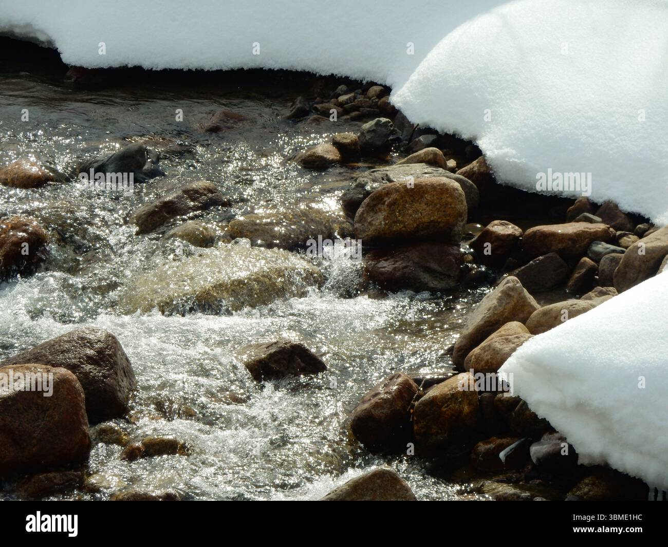 The sun reflects from a trickling stream with snow covered banks in ...