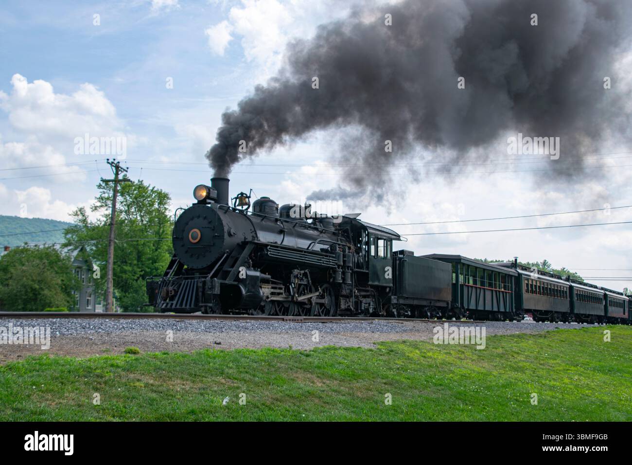 A historic narrow gauge steam locomotive moves along the tracks in a rural area, releasing black ...