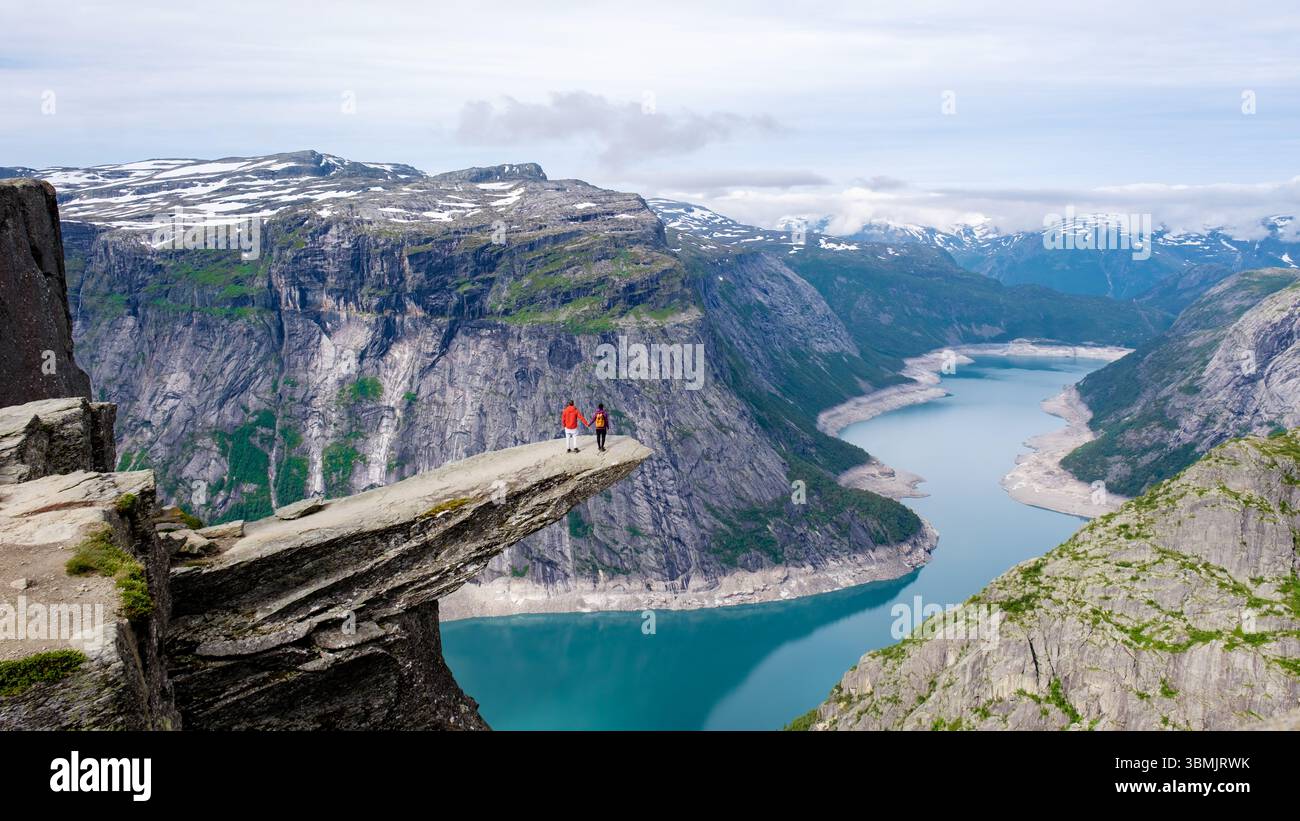 Two hikers stand on a dramatic cliff edge, gazing at the stunning fjord ...