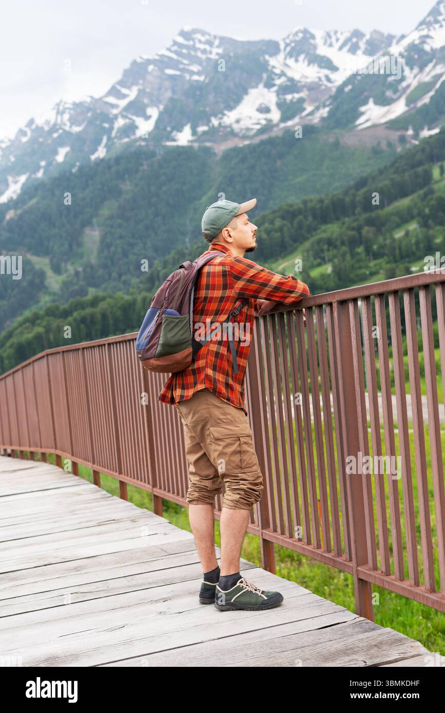 Male hiker in plaid shirt , backpack and cap leans on railing, admiring ...