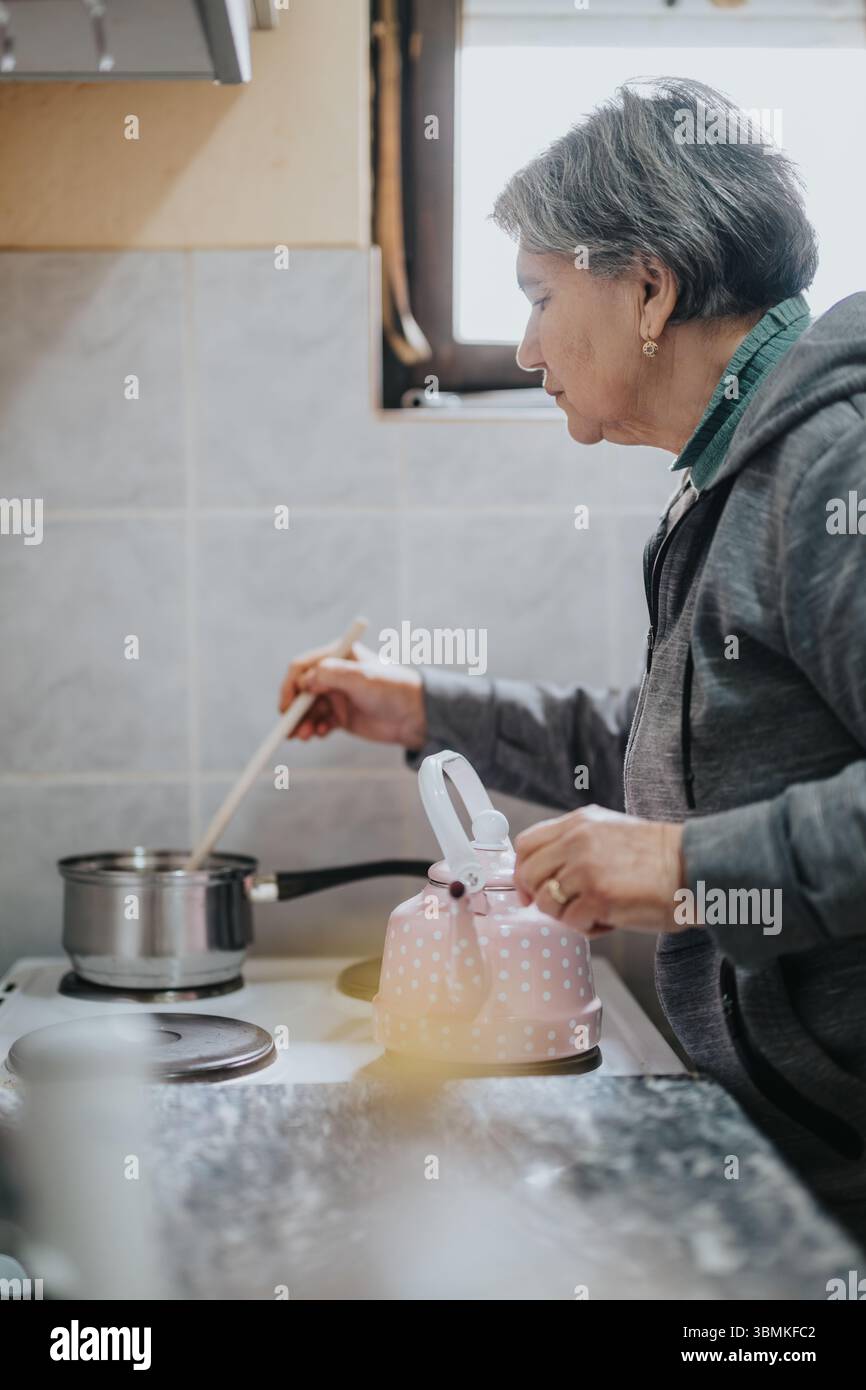 Elderly Woman Cooking on a Stove with Pink Teapot in a Bright Kitchen ...