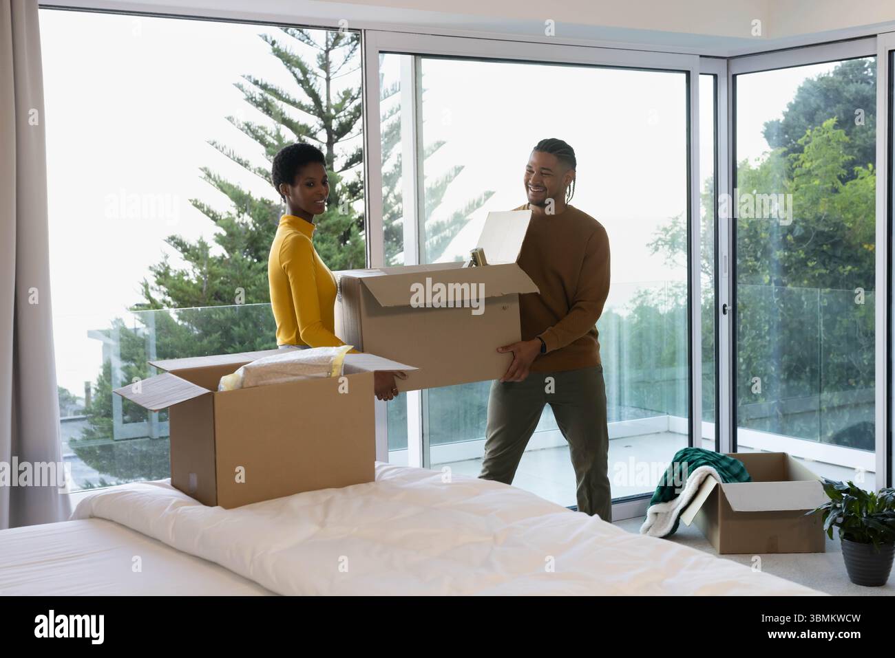 Diverse couple carrying large cardboard moving box in modern bedroom ...