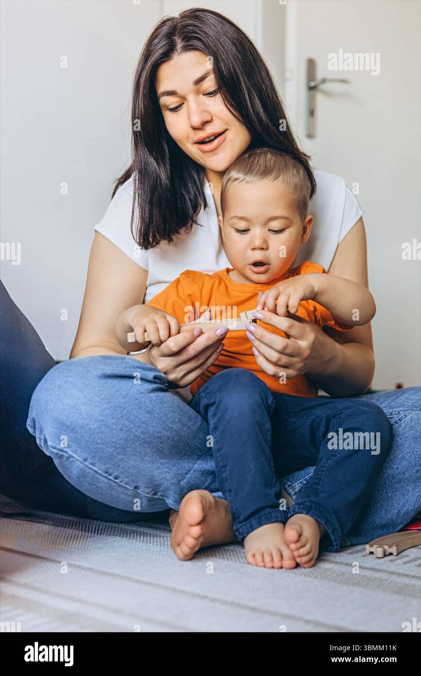 Loving mother sitting on the floor with her two-year-old son in a cozy playroom, smiling ...