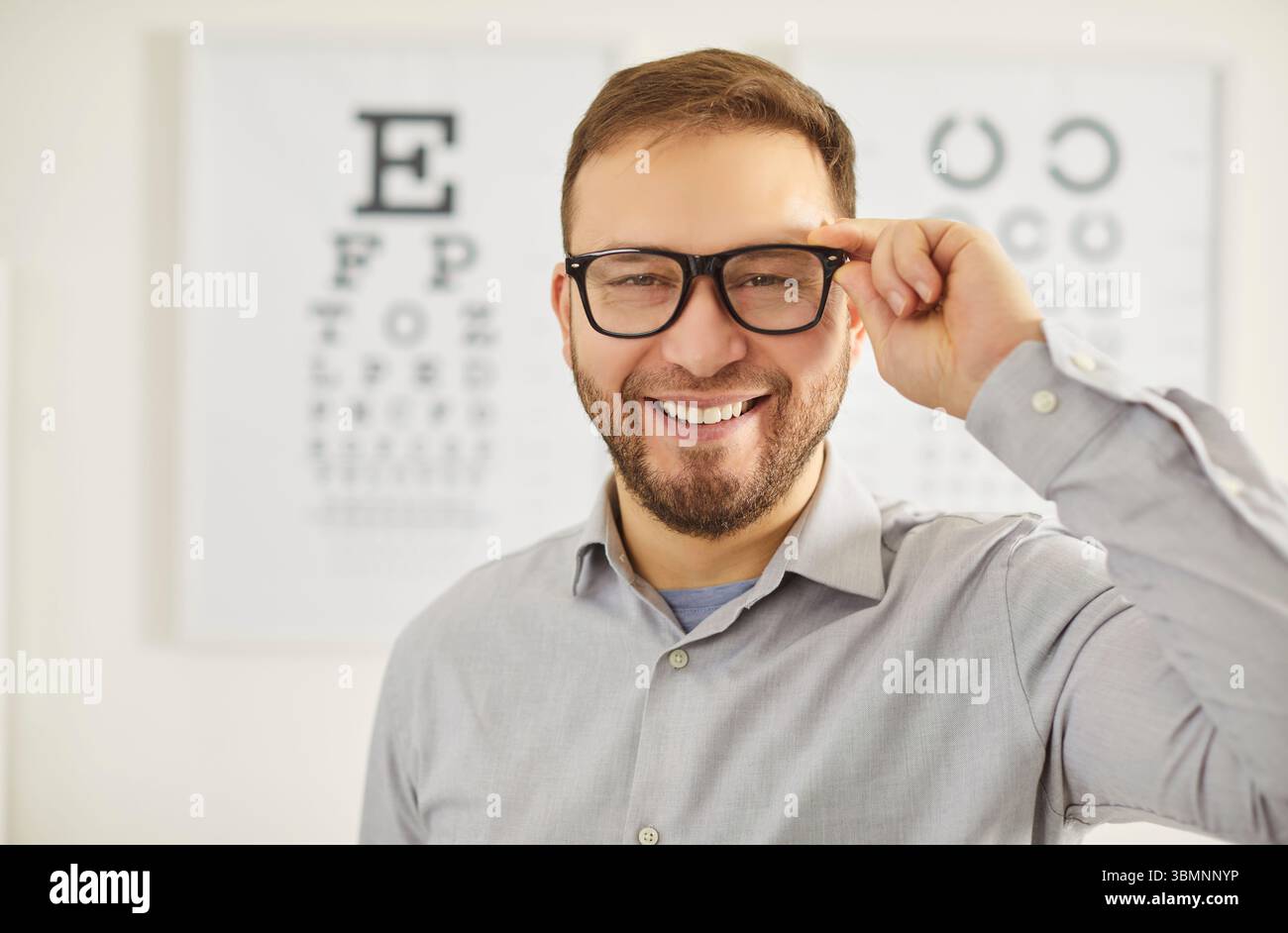 Man happy wearing glasses, posing near ophthalmologist eye chart visit ...