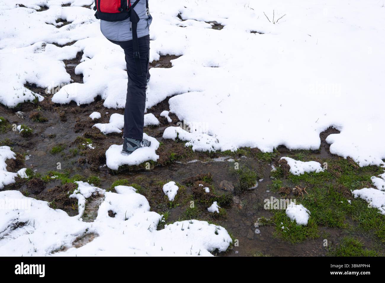A hiker steps on a patch of snow on a muddy stream in a winter ...