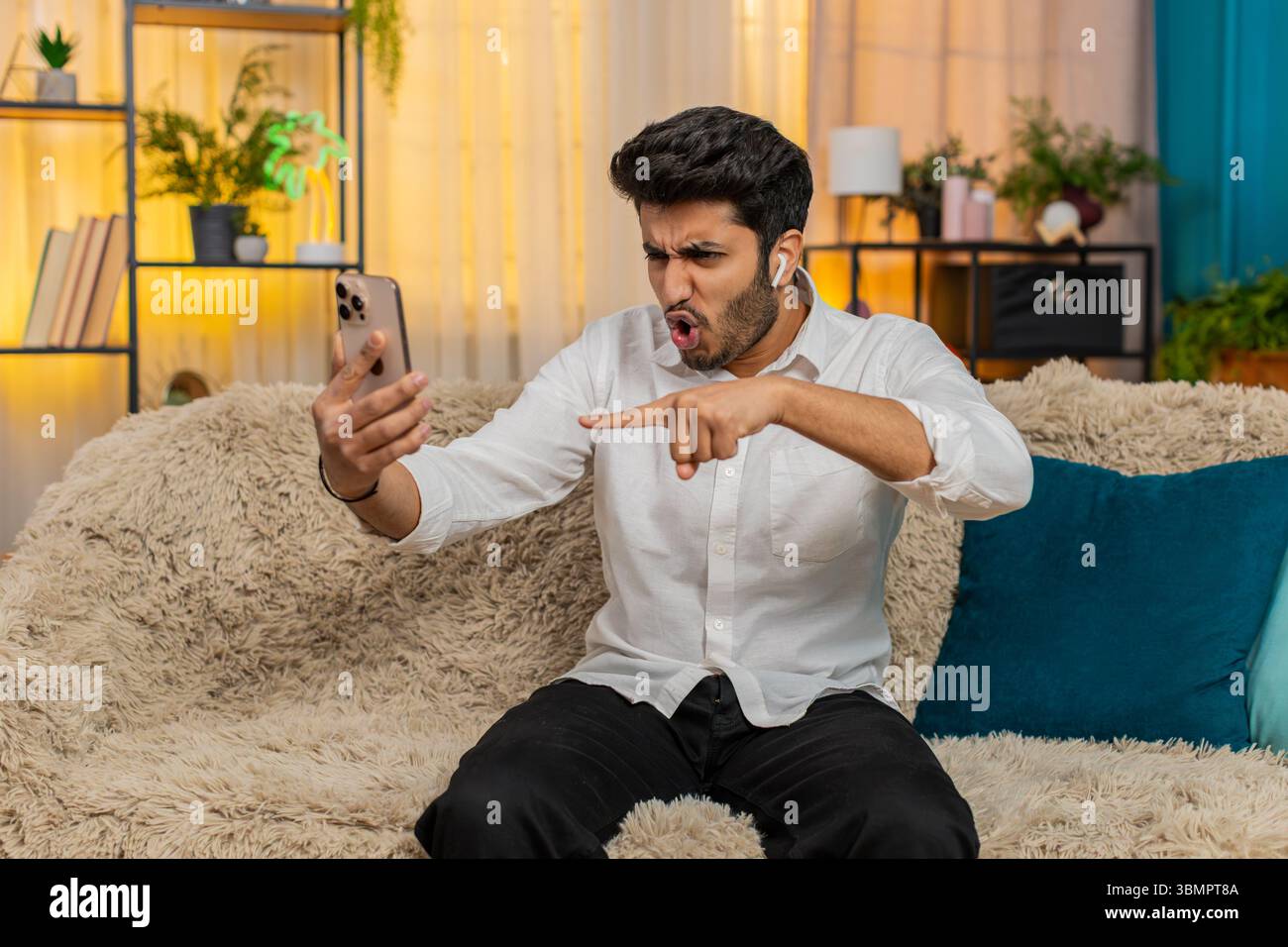 Indian man at home on sofa arguing during a video call, speaking through earphones. Arabian guy ...