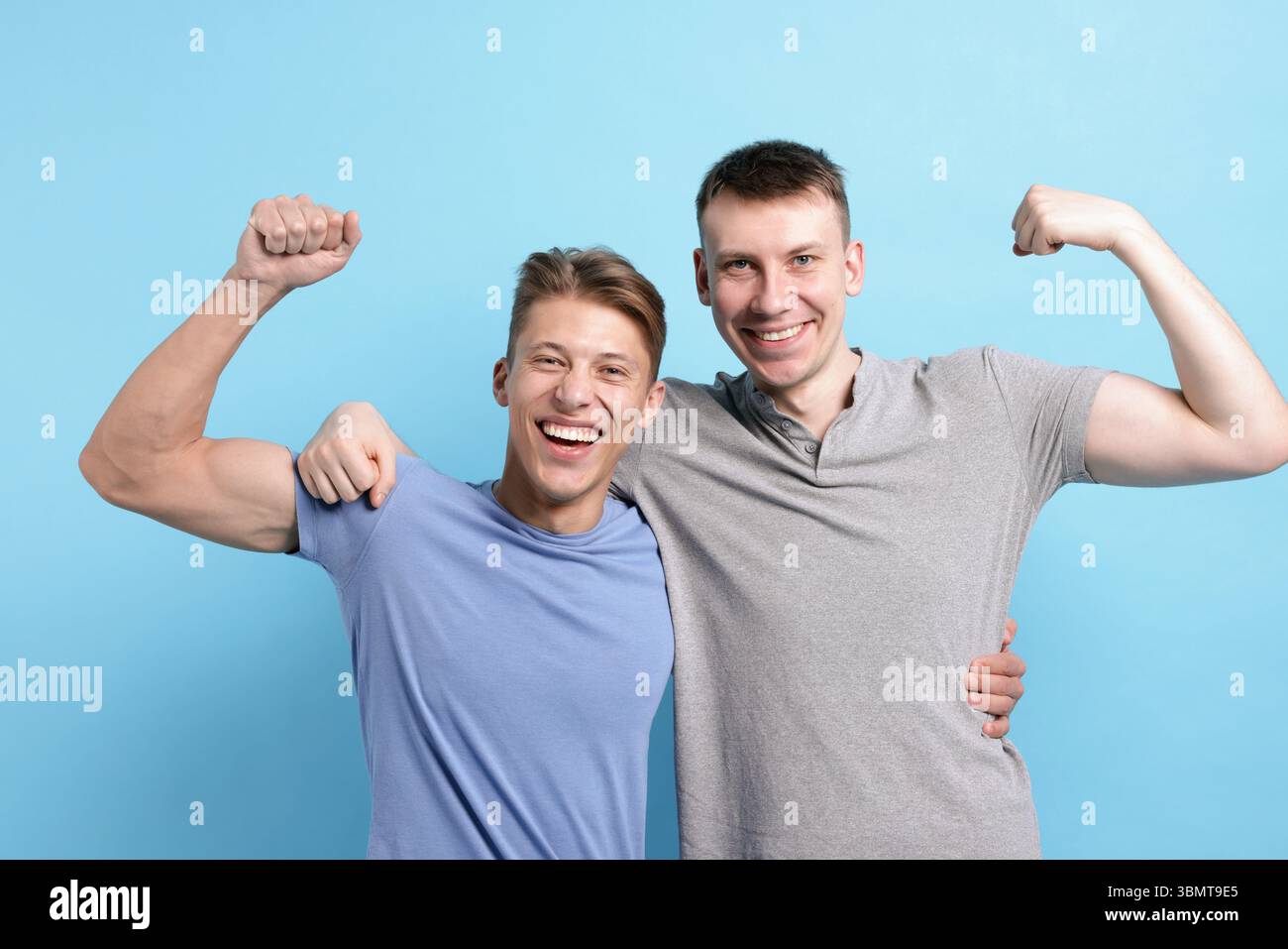 Two friends hugging and showing their biceps on light blue background ...