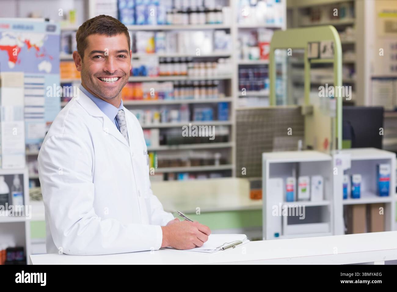 Male pharmacist in lab coat writing on clipboard behind counter with ...