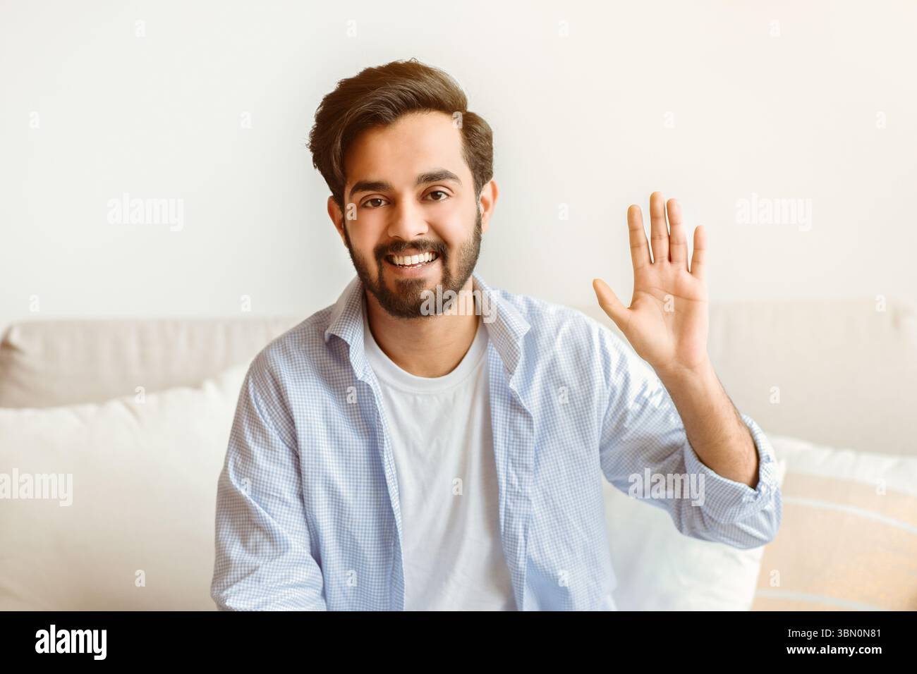 Greeting. Friendly eastern man waving hello while sitting on couch at ...