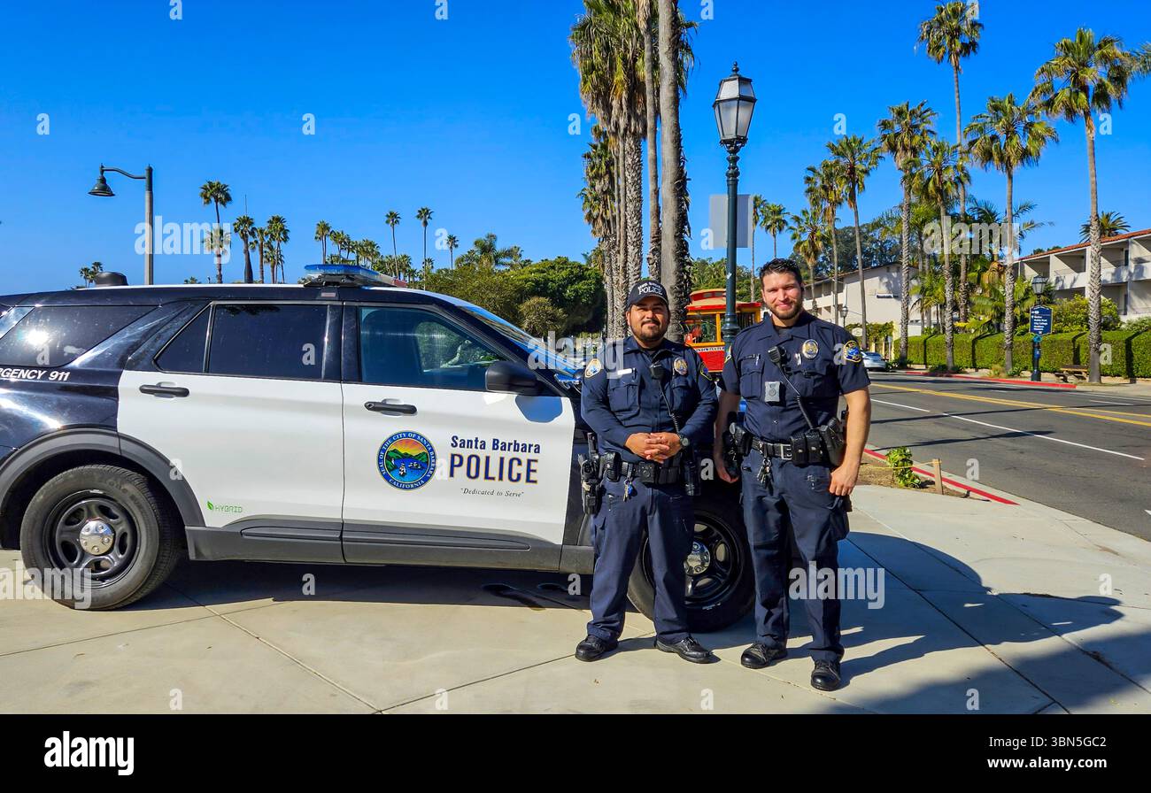 Two smiling police officers pose beside their patrol car in sunny Santa ...
