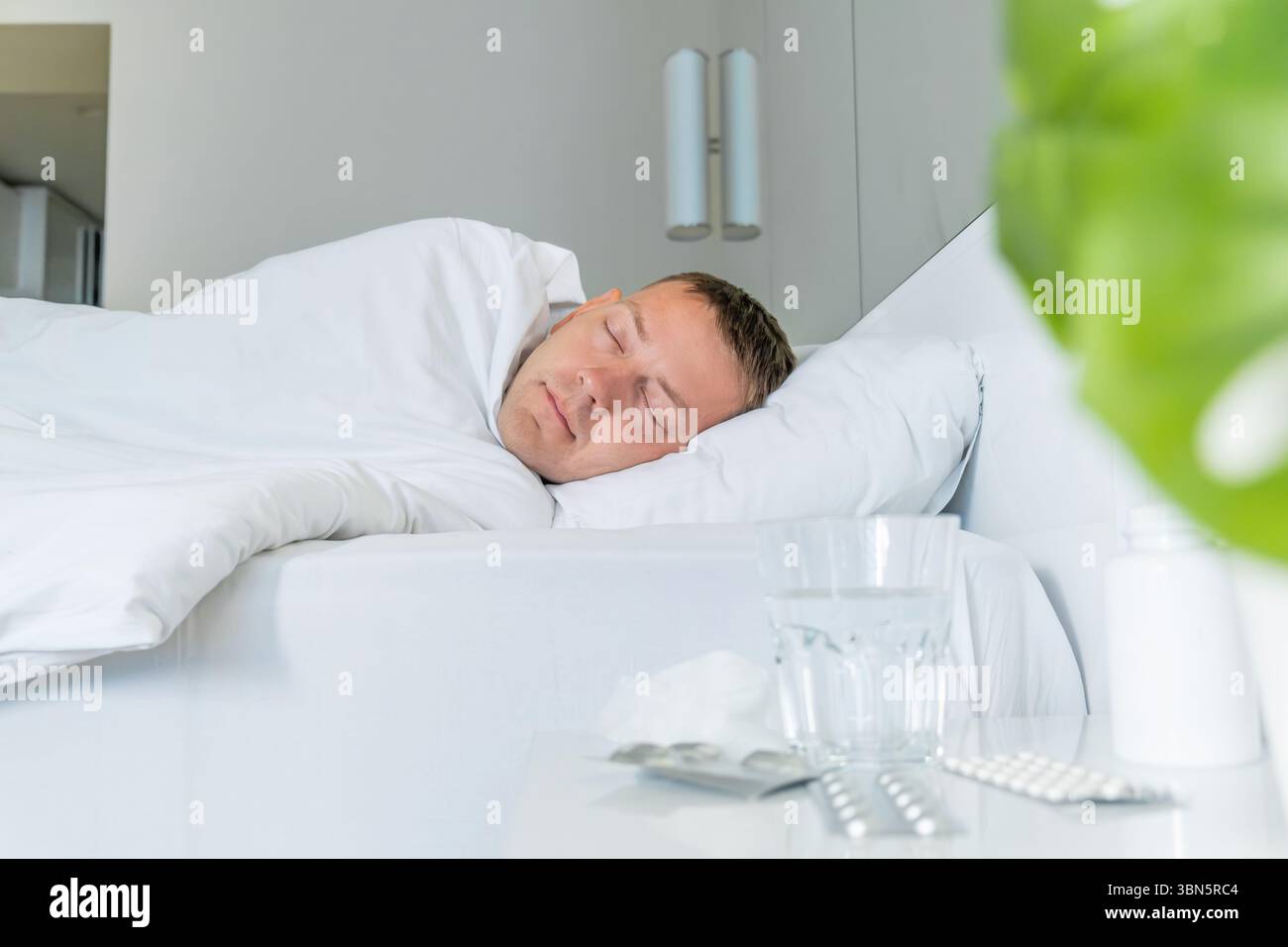 Sick man sleeping in bed under blanket with glass of water and medicine ...
