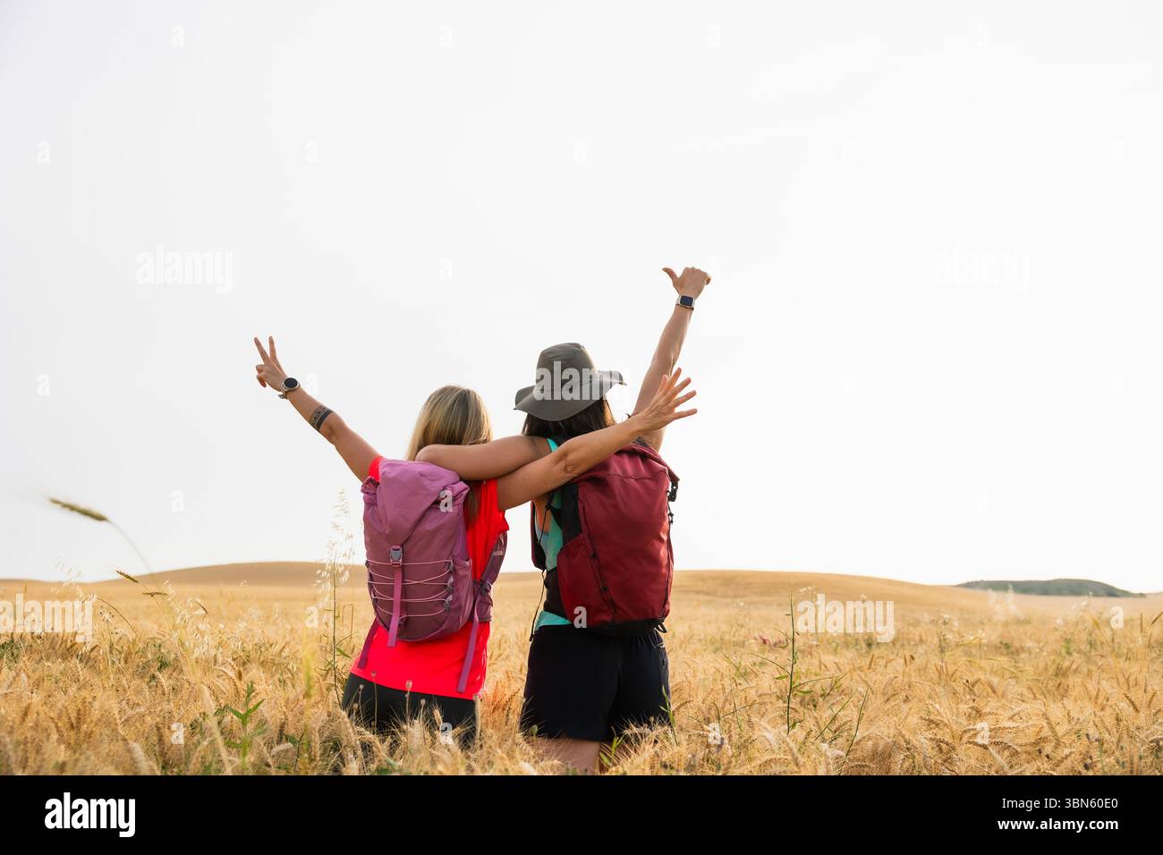 Two female hikers with backpacks stand arm in arm in a golden wheat ...