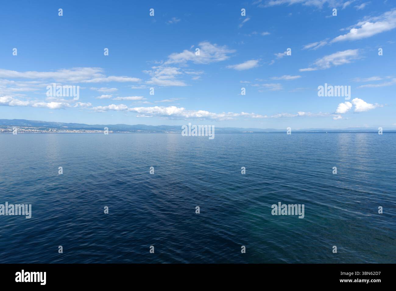 A tranquil blue sea stretches under a vast sky dotted with white clouds ...