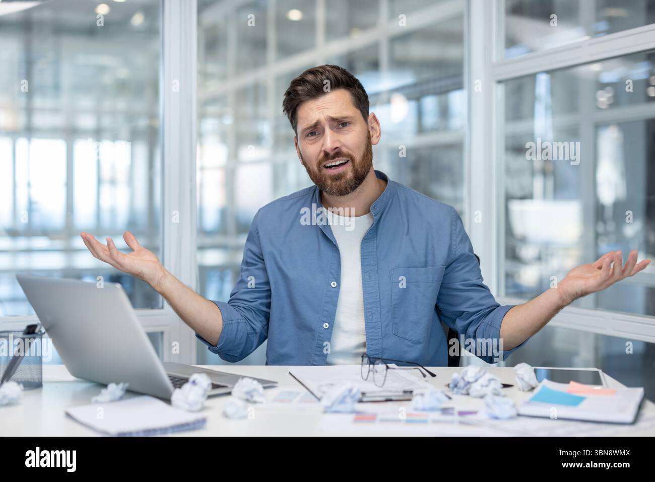 A frustrated man sits at a desk strewn with crumpled paper, appearing ...